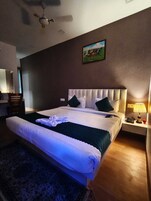 Quarto de hotel com uma cama grande, ventoinha no teto, mesa de cabeceira com abajur e um quadro na parede.
