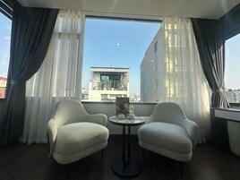 Quarto com duas poltronas brancas e uma pequena mesa redonda, janela com cortinas leves e vista de um edifício na rua.