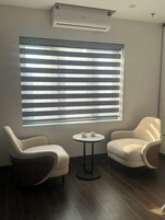 Quarto moderno com uma janela grande coberta por persianas horizontais, duas poltronas brancas e uma pequena mesa redonda com um bule e xícara.