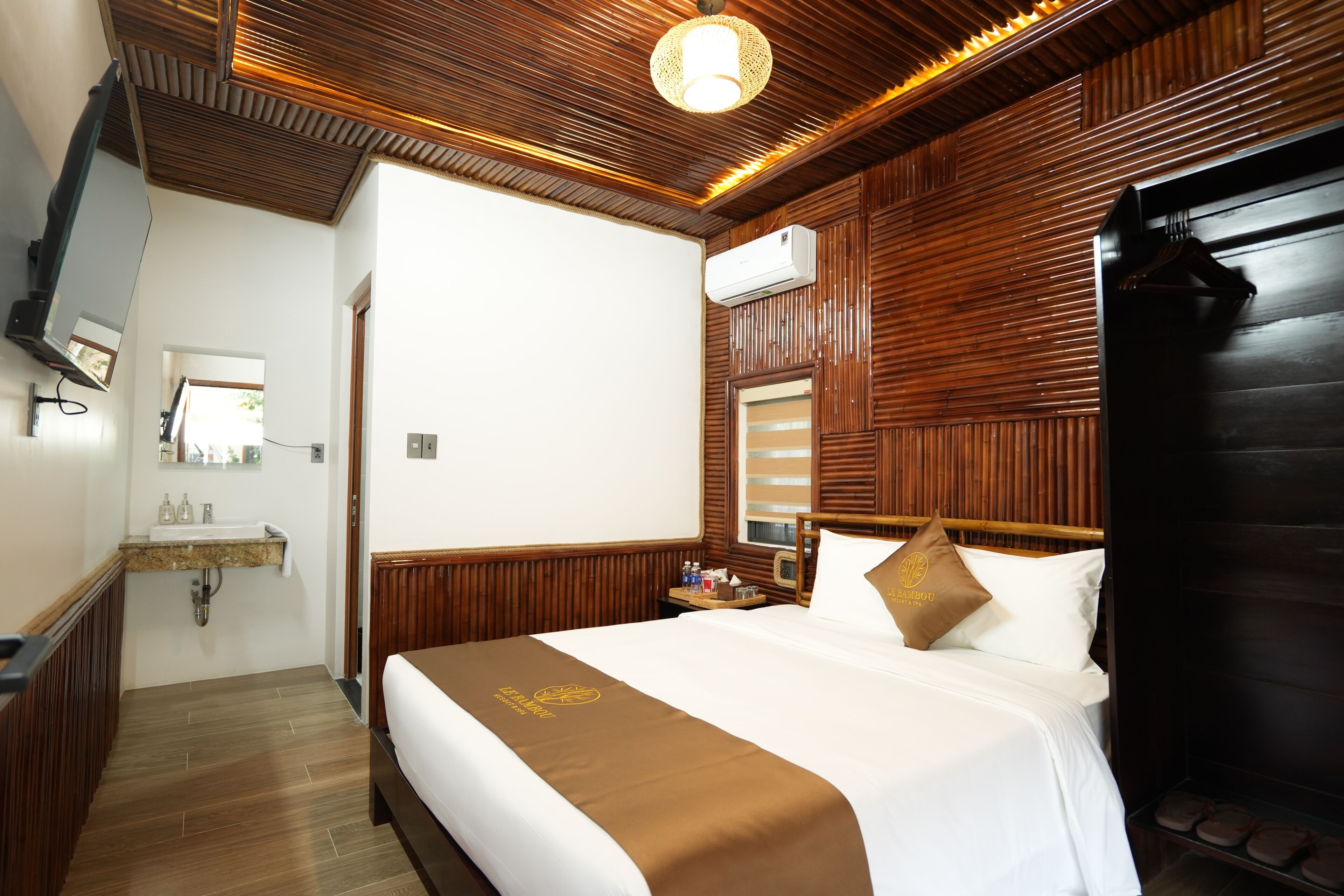 Quarto de hotel com teto de madeira, cama com colcha marrom, mesa de cabeceira com abajur e espelho sobre a pia.