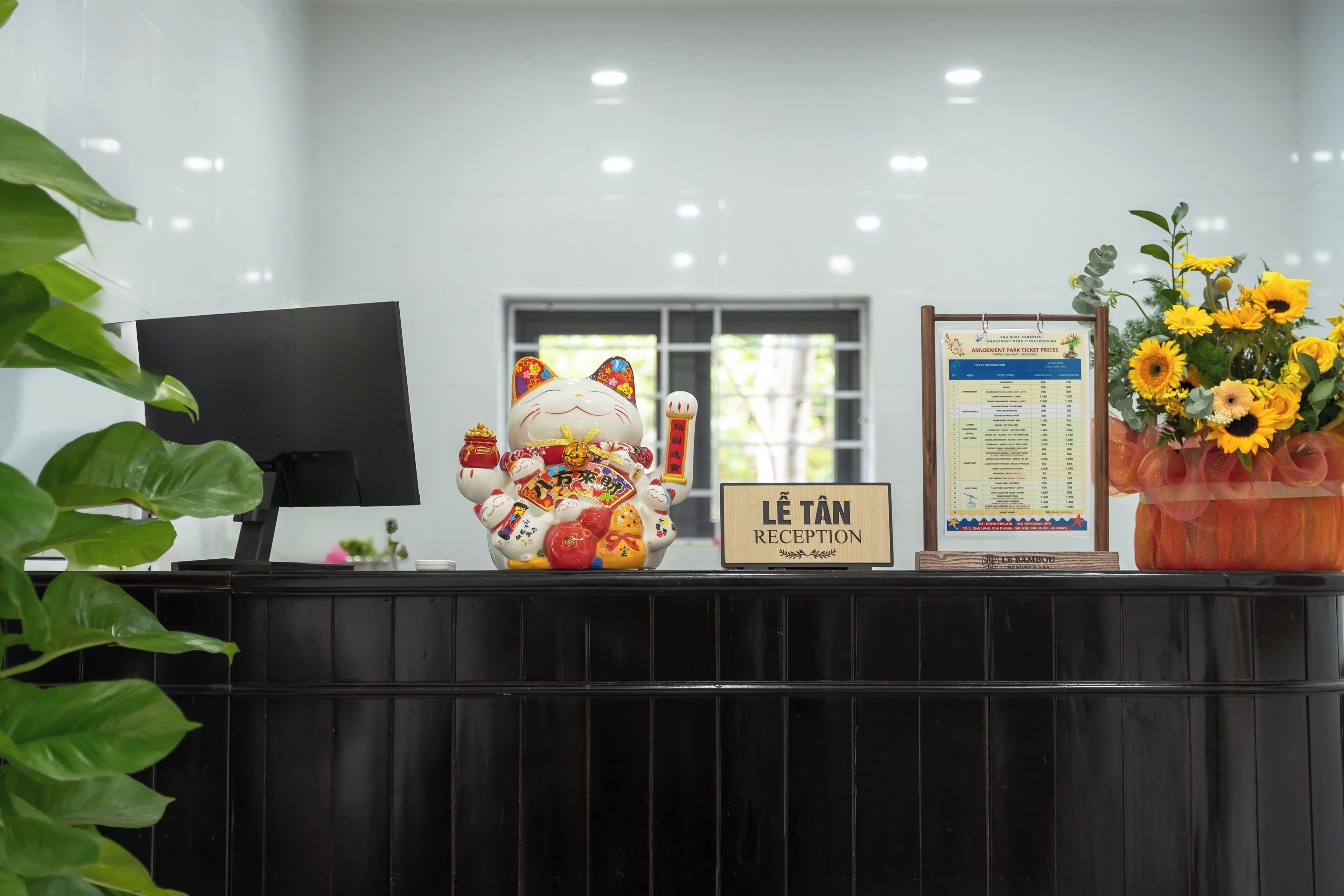 Uma mesa de recepção com um monitor, uma estatueta de gato decorativa, uma placa com a inscrição "LE TÃN RECEPTION" e um cardápio emoldurado.