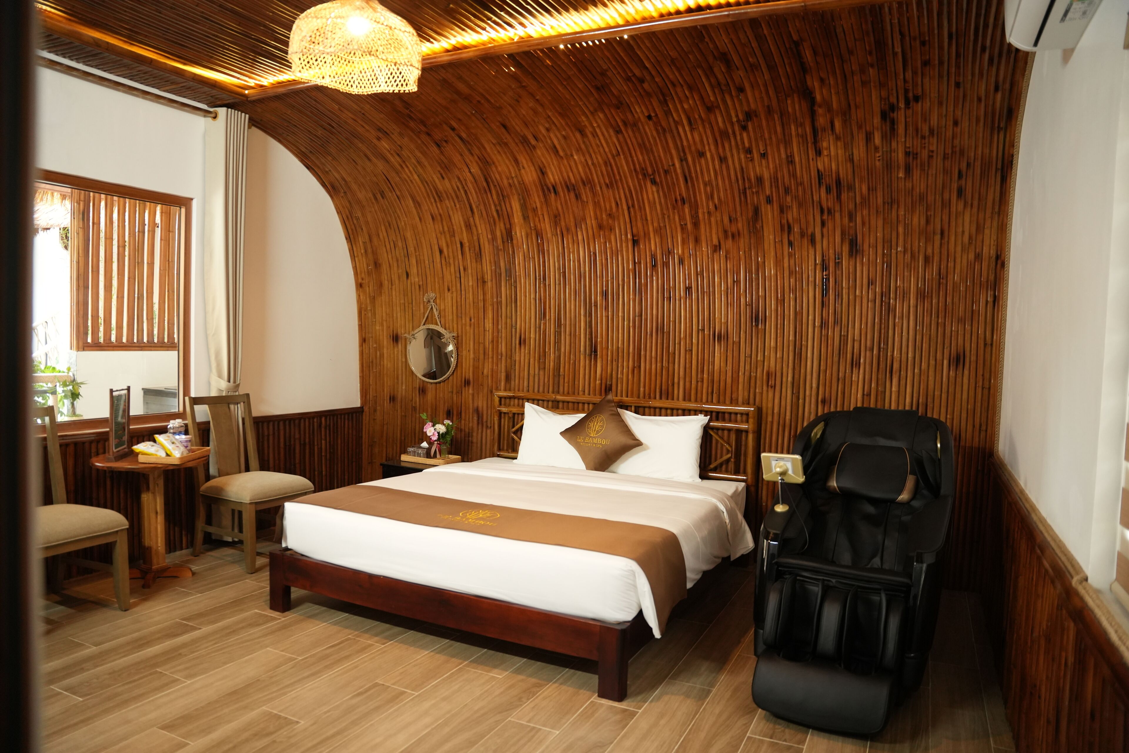 Quarto de hotel com teto de madeira, cama com colcha marrom, cadeira de massagem e uma pequena mesa com abajur.