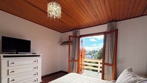 Quarto com cama, penteadeira, televisão por cima e varanda com cortinas.