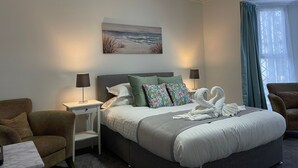 Quarto de hotel com cama, duas mesas de cabeceira, abajur, cadeira, quadro e janela com cortinas.