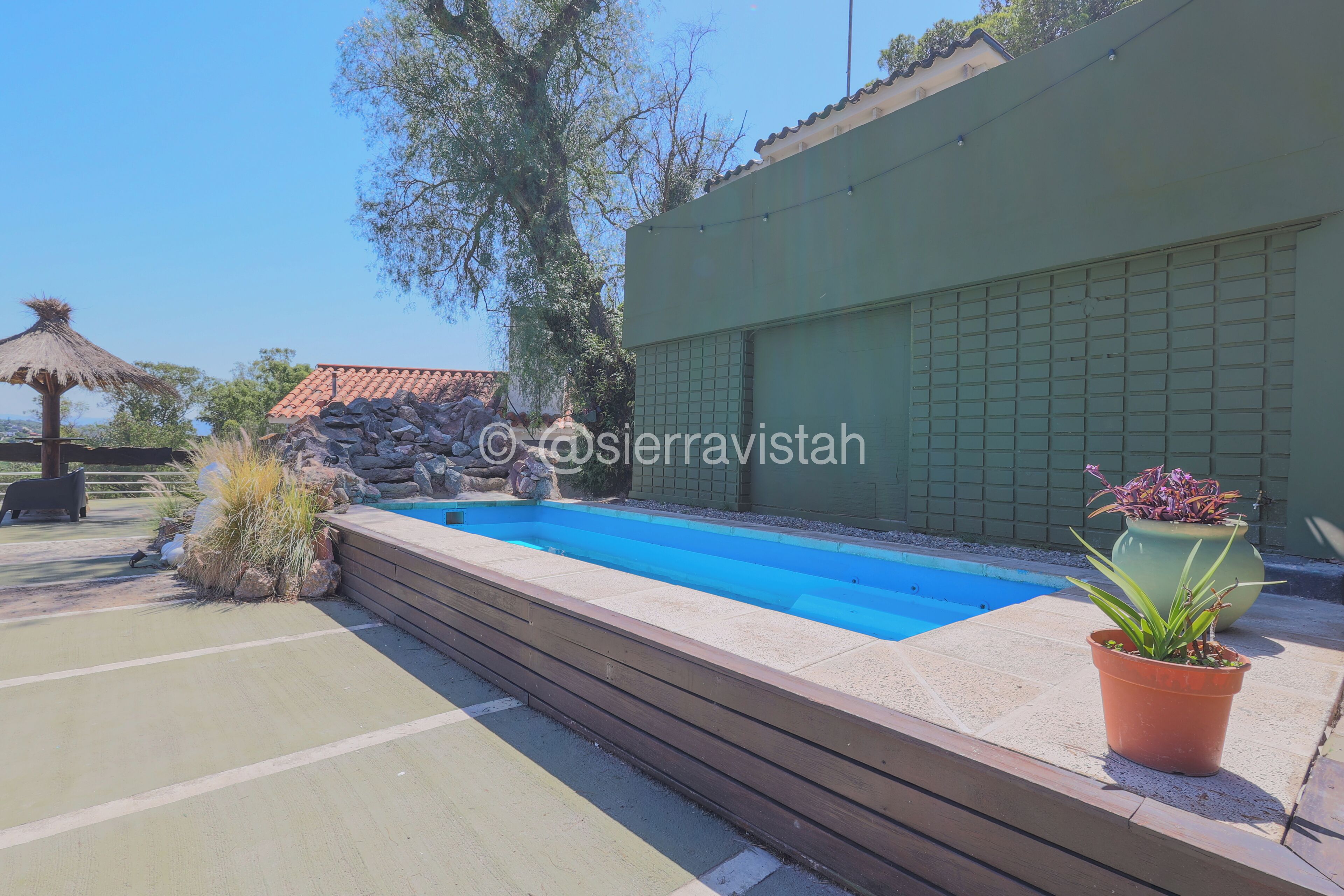 Uma piscina com deck de madeira, parede de pedra e um edifício verde ao fundo.