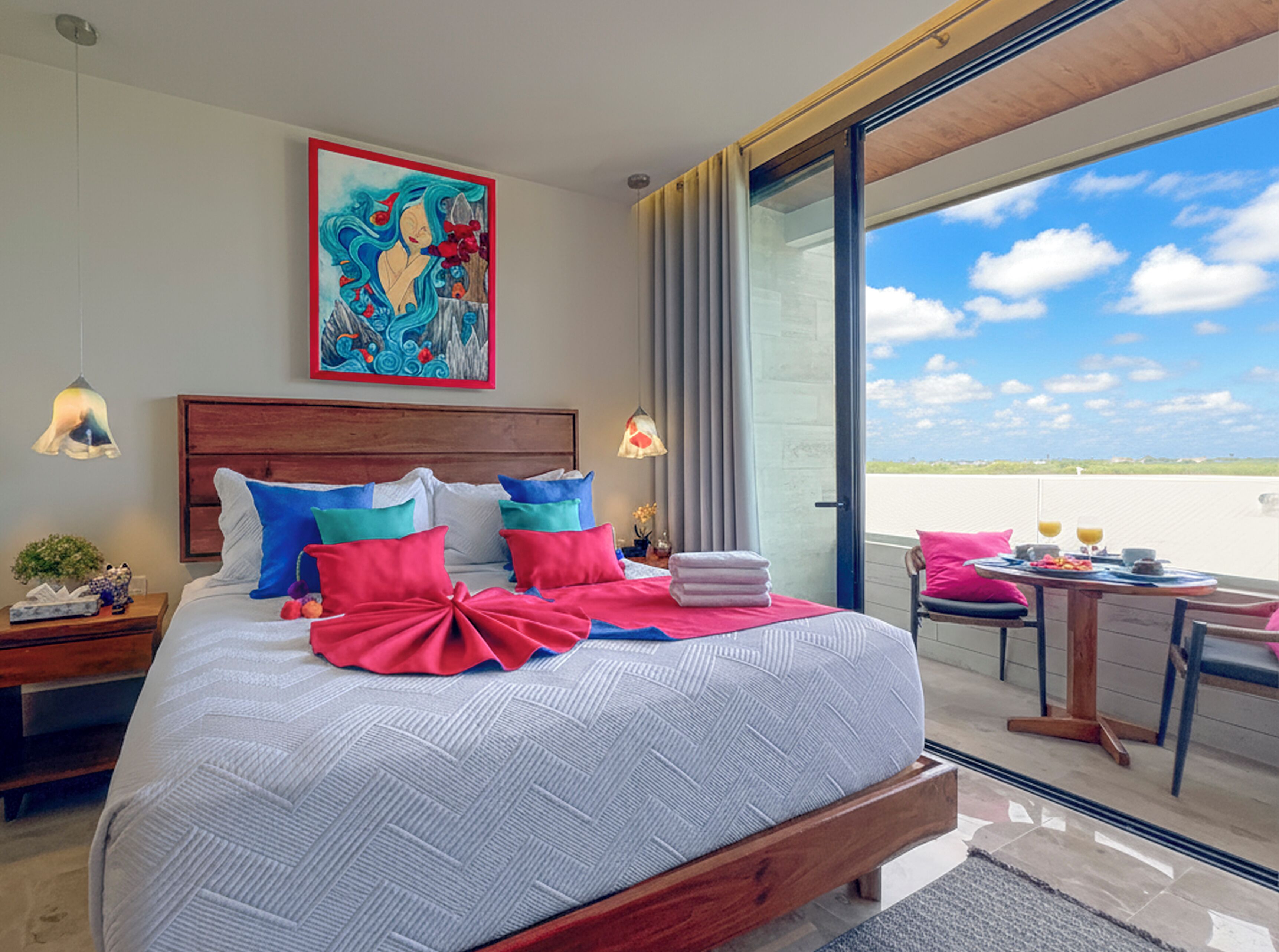 Quarto moderno com cama grande, almofadas coloridas, um quadro na parede e uma varanda com mesa e cadeiras.