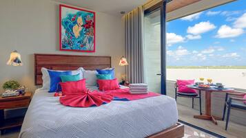 Quarto moderno com cama grande, almofadas coloridas, um quadro na parede e uma varanda com mesa e cadeiras.