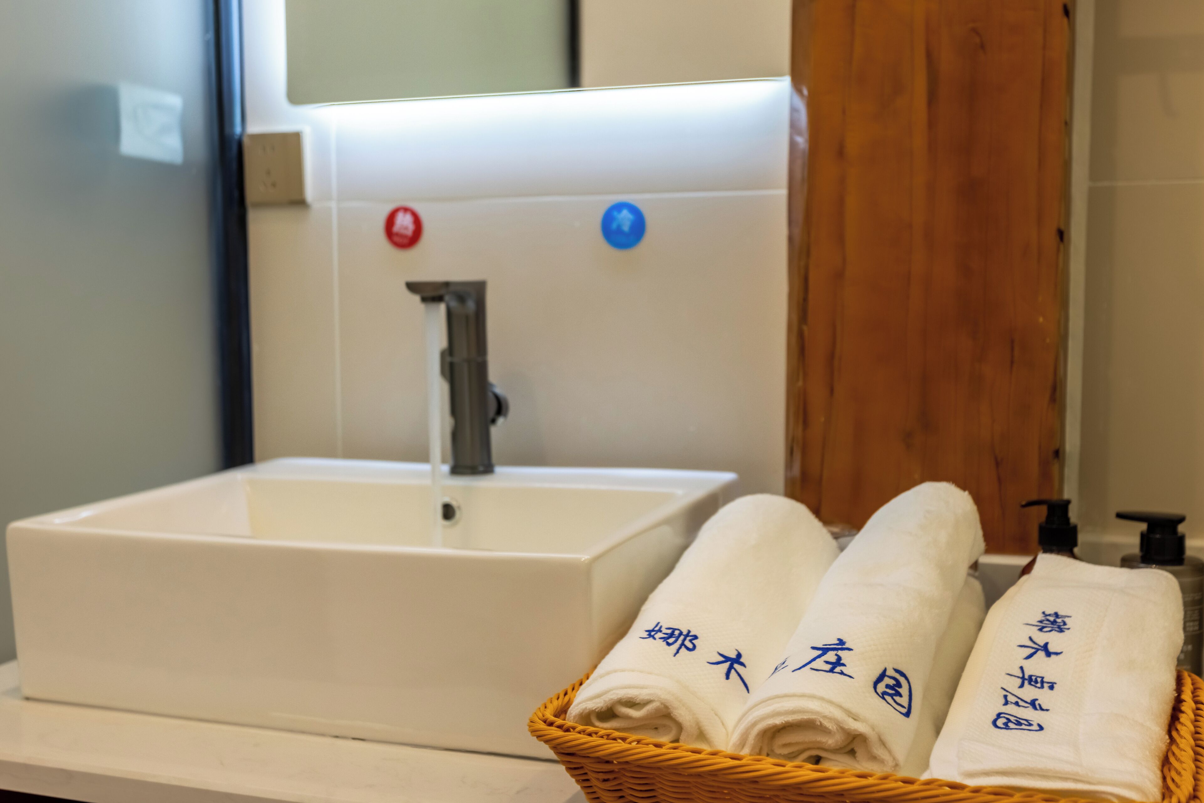 Uma casa de banho com uma pia branca, um espelho e uma porta de madeira. Há três toalhas com caracteres japoneses.