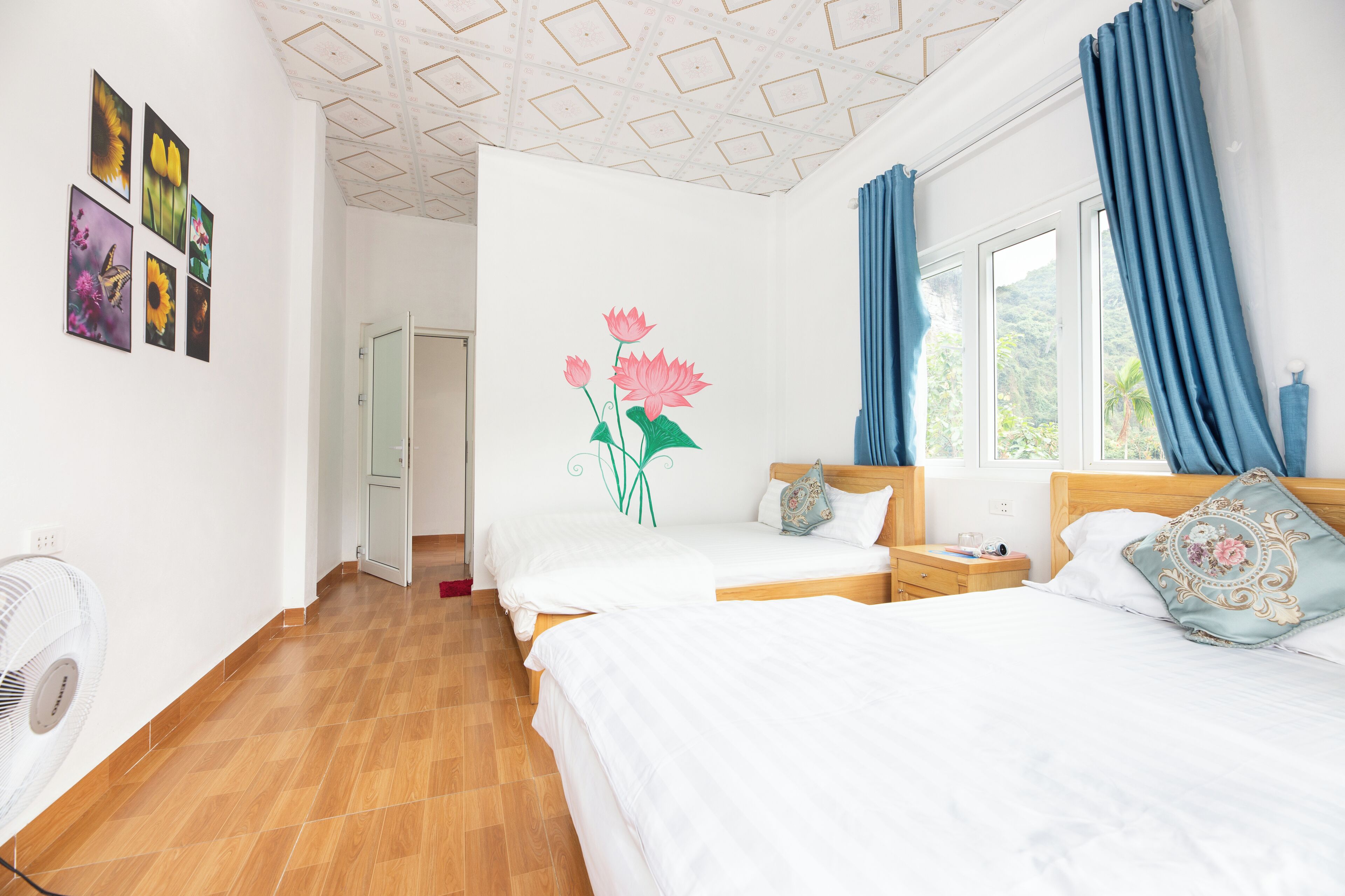 Quarto de hotel com duas camas, um ventilador de teto, uma janela com cortinas e obras de arte na parede.