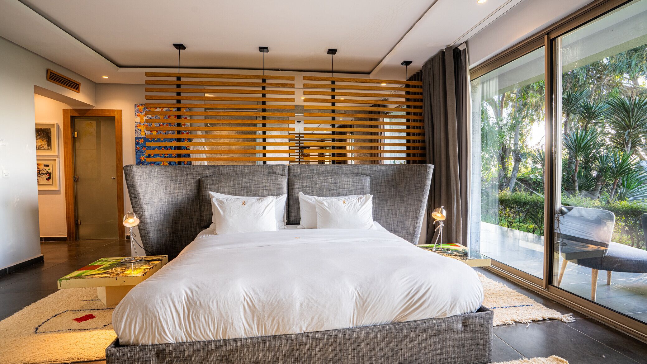 Quarto moderno com uma cama grande, cabeceira de ripas de madeira e vista para o verde através de grandes janelas.