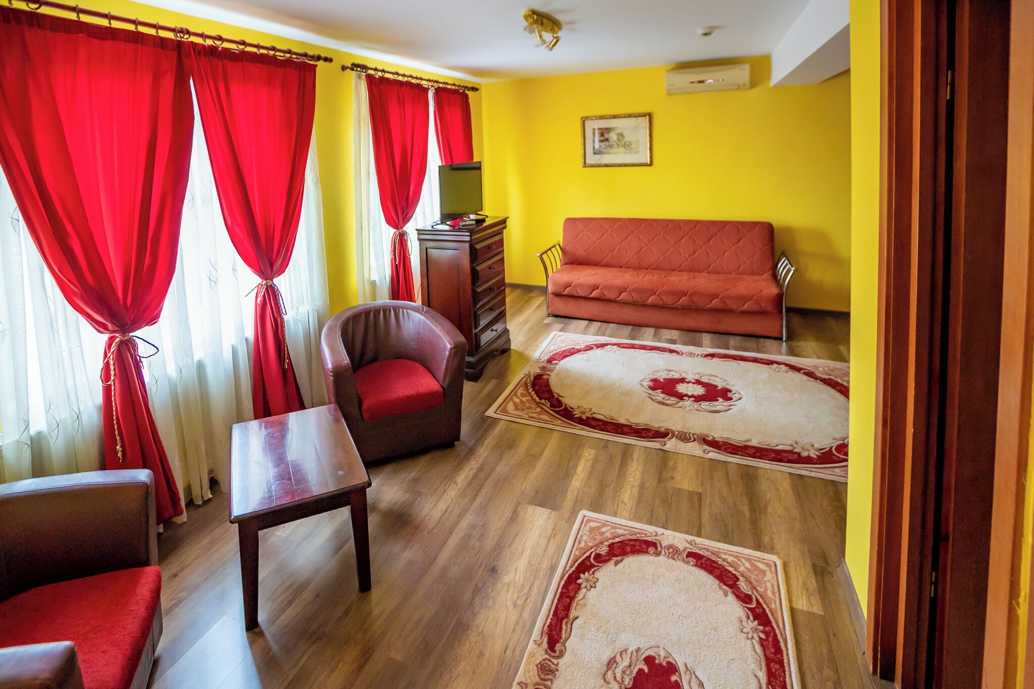 Quarto com sofá vermelho, mesa de madeira e cadeira vermelha. Há uma TV em um armário de madeira e uma cortina vermelha presa na parte inferior da janela.