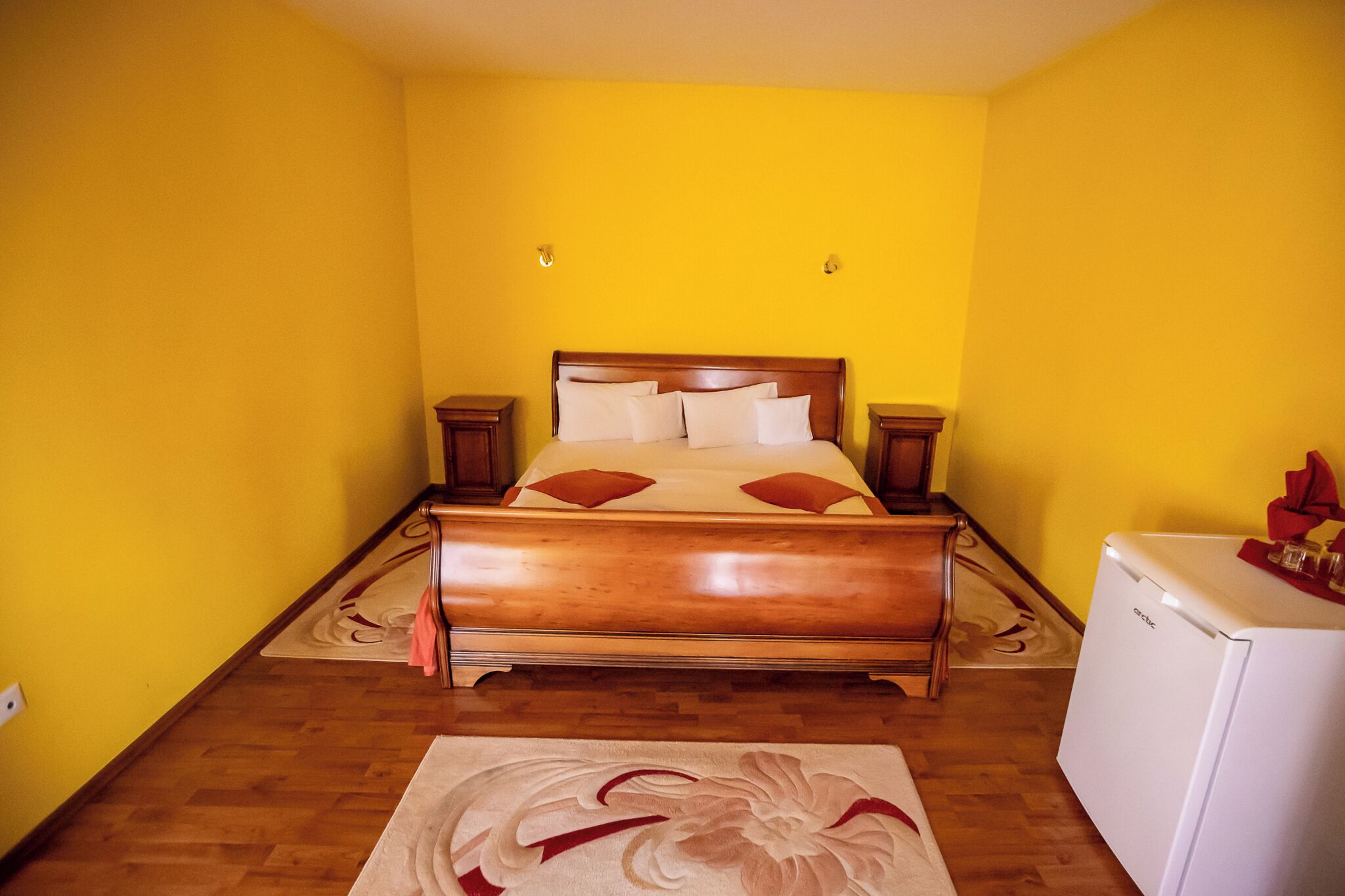 Quarto com cama de madeira, paredes amarelas e eletrodoméstico branco.