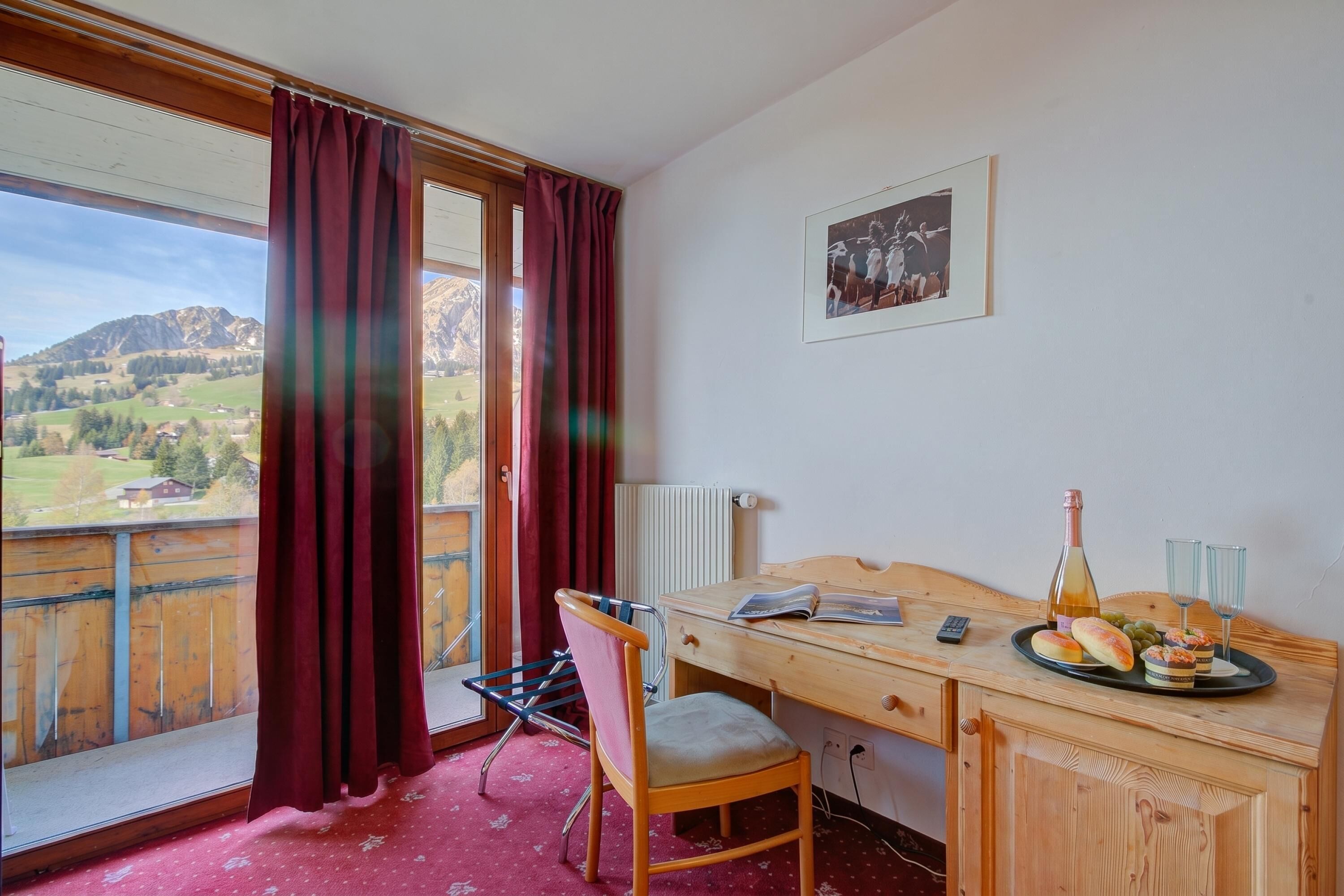 Um quarto com escrivaninha de madeira, uma cadeira, uma mesa de jantar com comida, uma garrafa, copos, um radiador e uma janela com cortinas.