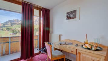 Um quarto com escrivaninha de madeira, uma cadeira, uma mesa de jantar com comida, uma garrafa, copos, um radiador e uma janela com cortinas.