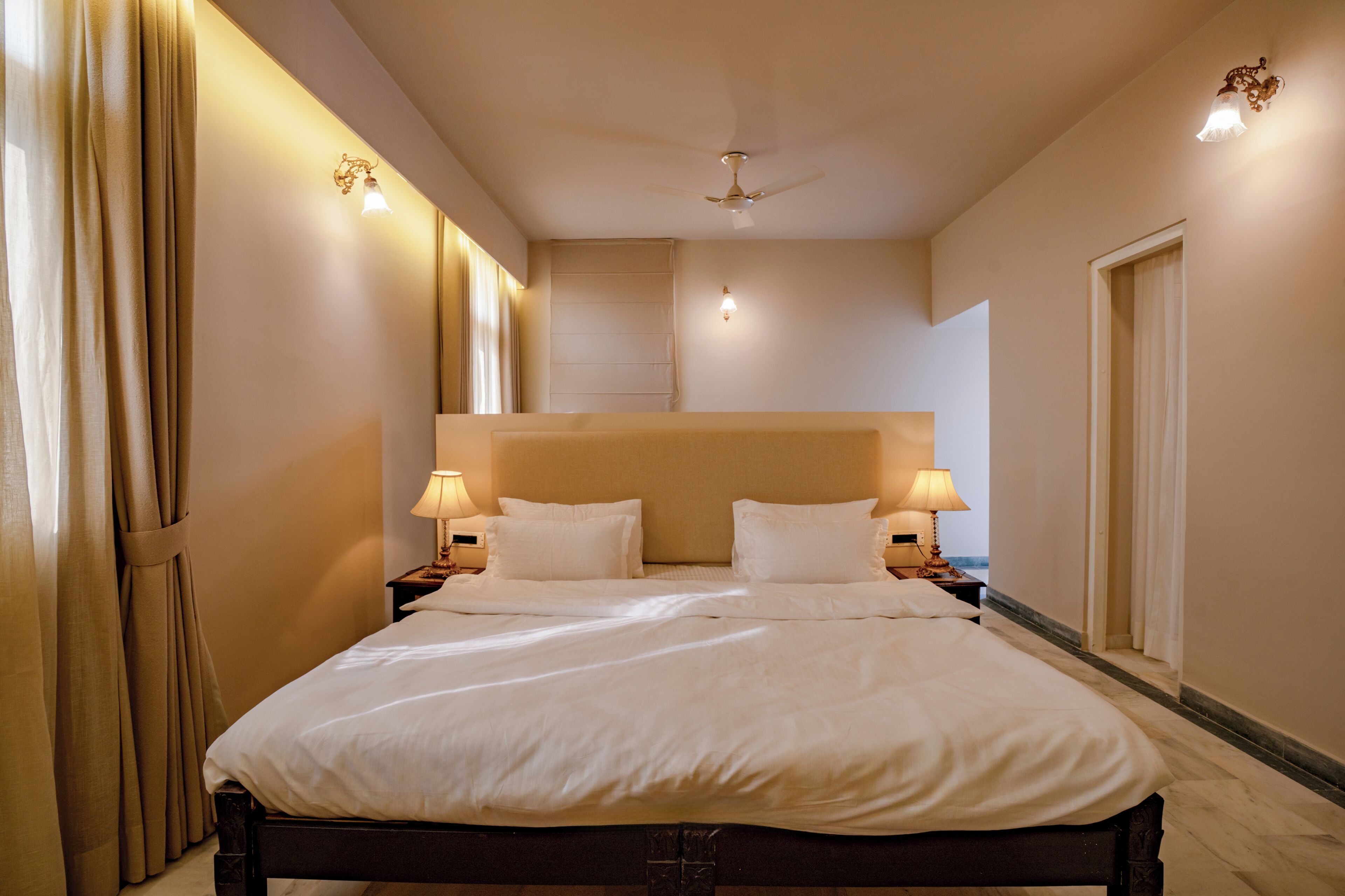 Quarto de hotel com uma cama grande, duas mesas de cabeceira com abajures, um ventilador de teto e vista para o mar pela janela.