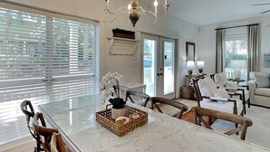 Uma sala de jantar com mesa de vidro, cadeiras, um lustre e vista através de grandes janelas.