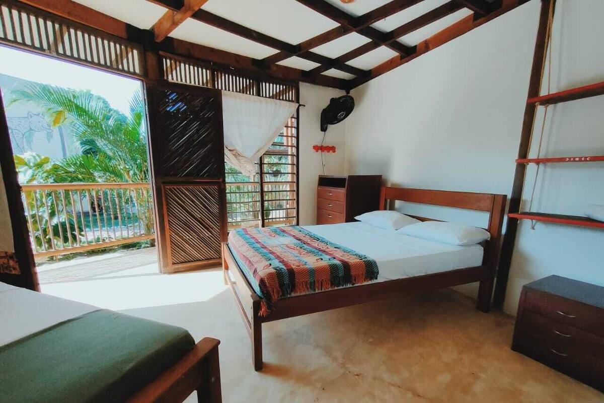 Quarto com cama, mesa de cabeceira, cômoda e varanda com vista para o verde.