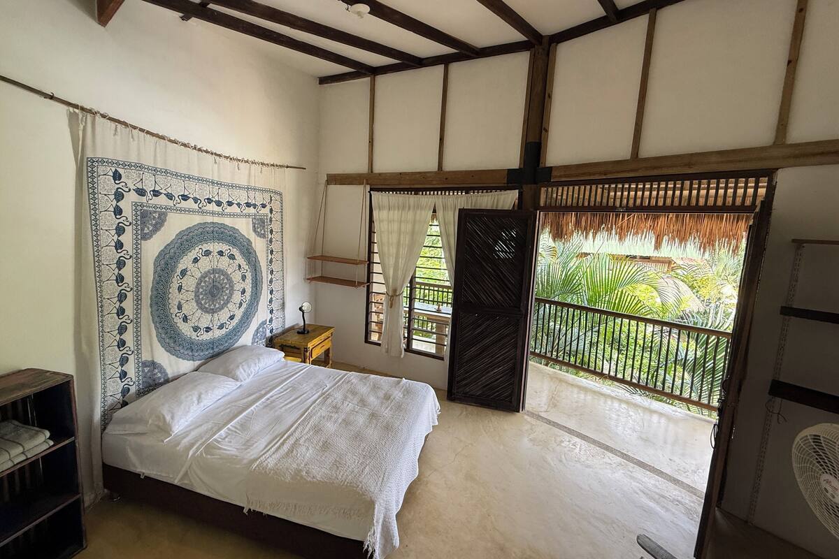 Quarto com cama, mesa de cabeceira, luminária pendente, janela com vista para o verde e varanda com guarda-corpo.