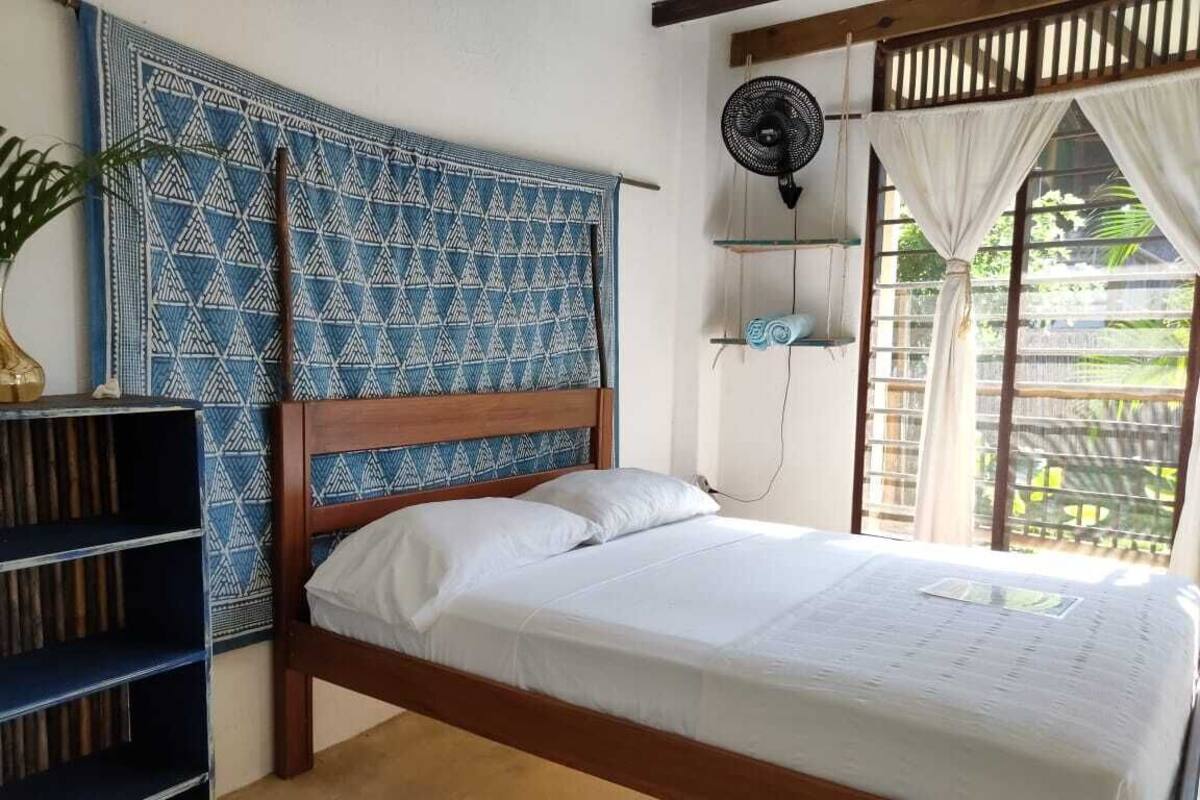 Uma cama de solteiro com cabeceira de madeira, um ventilador na mesinha de cabeceira e uma tapeçaria estampada na parede.