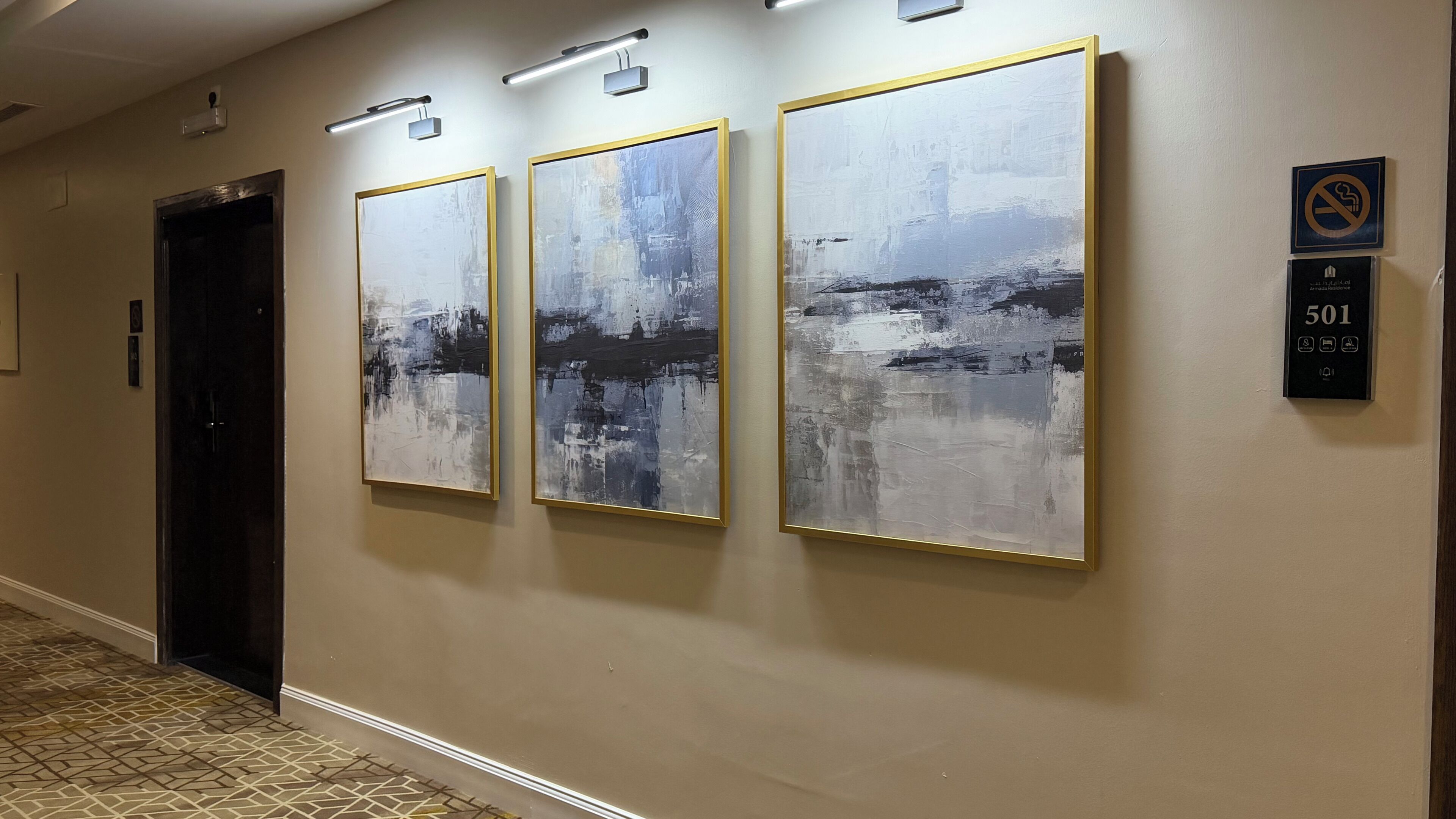 Um corredor com três quadros abstratos, uma porta, um aviso de proibição de fumar e uma placa com o número do quarto.