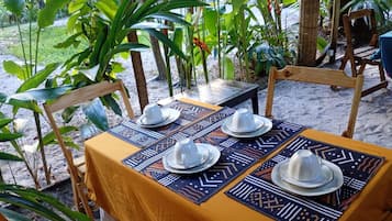 Mesa posta com pratos e xícaras brancos, cercada por cadeiras, em um jardim com diversas plantas.