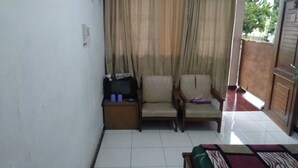 Quarto pequeno e bem iluminado com cama, duas cadeiras, mesa de cabeceira e janela com cortinas.