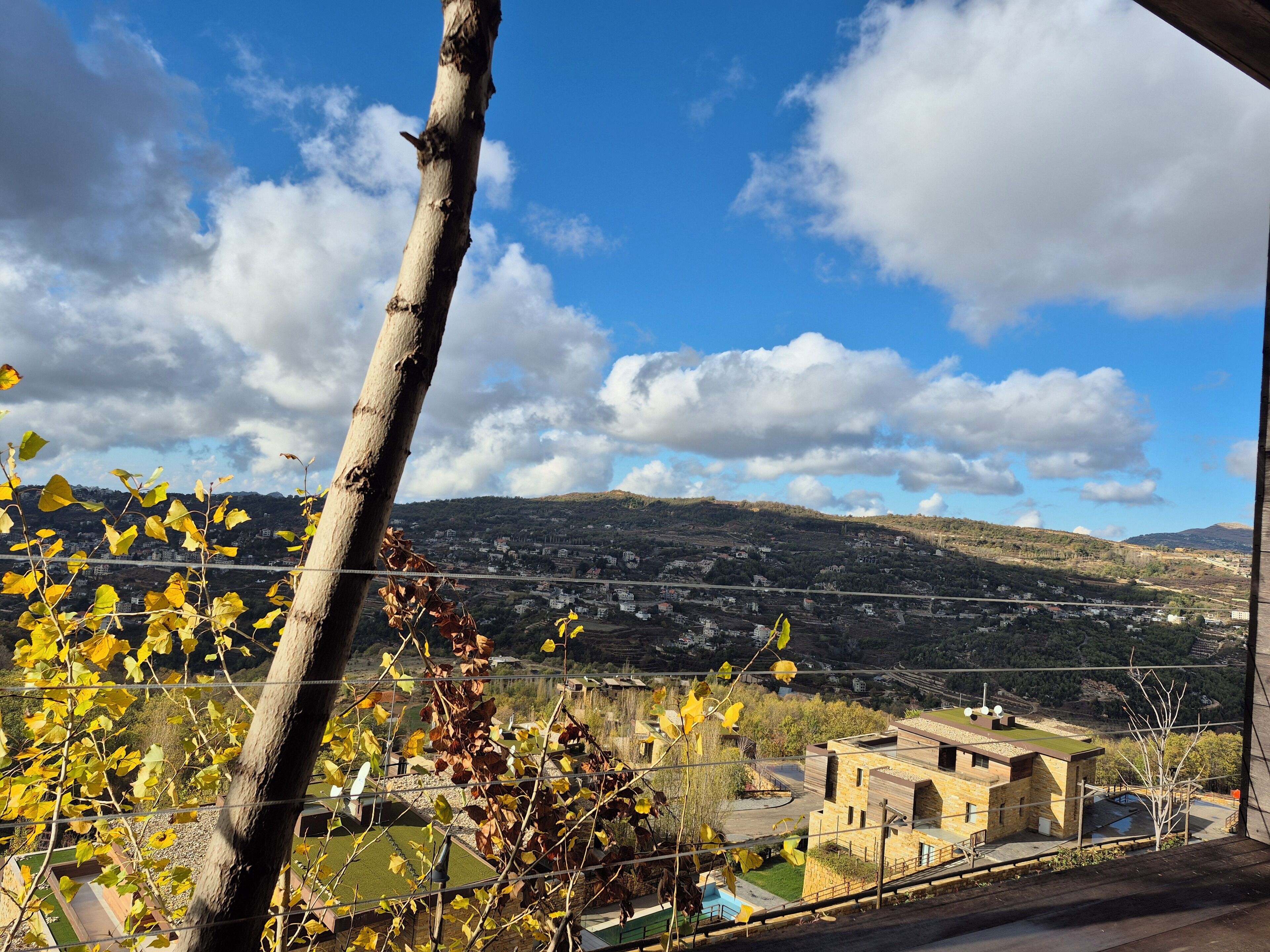 Vista de uma varanda, com um galho de árvore em primeiro plano, céu azul límpido e paisagem distante com edifícios e colinas.