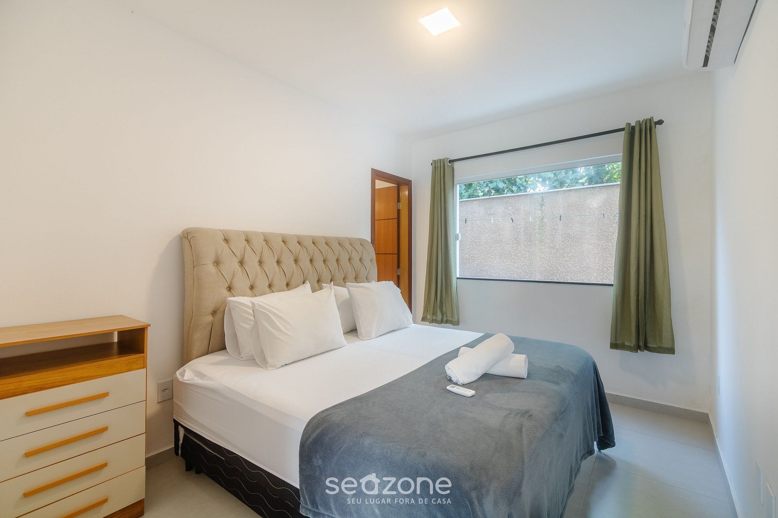 Uma cama bem arrumada com um cobertor cinzento, um penteado de madeira com gavetas e uma janela com cortinas verdes.