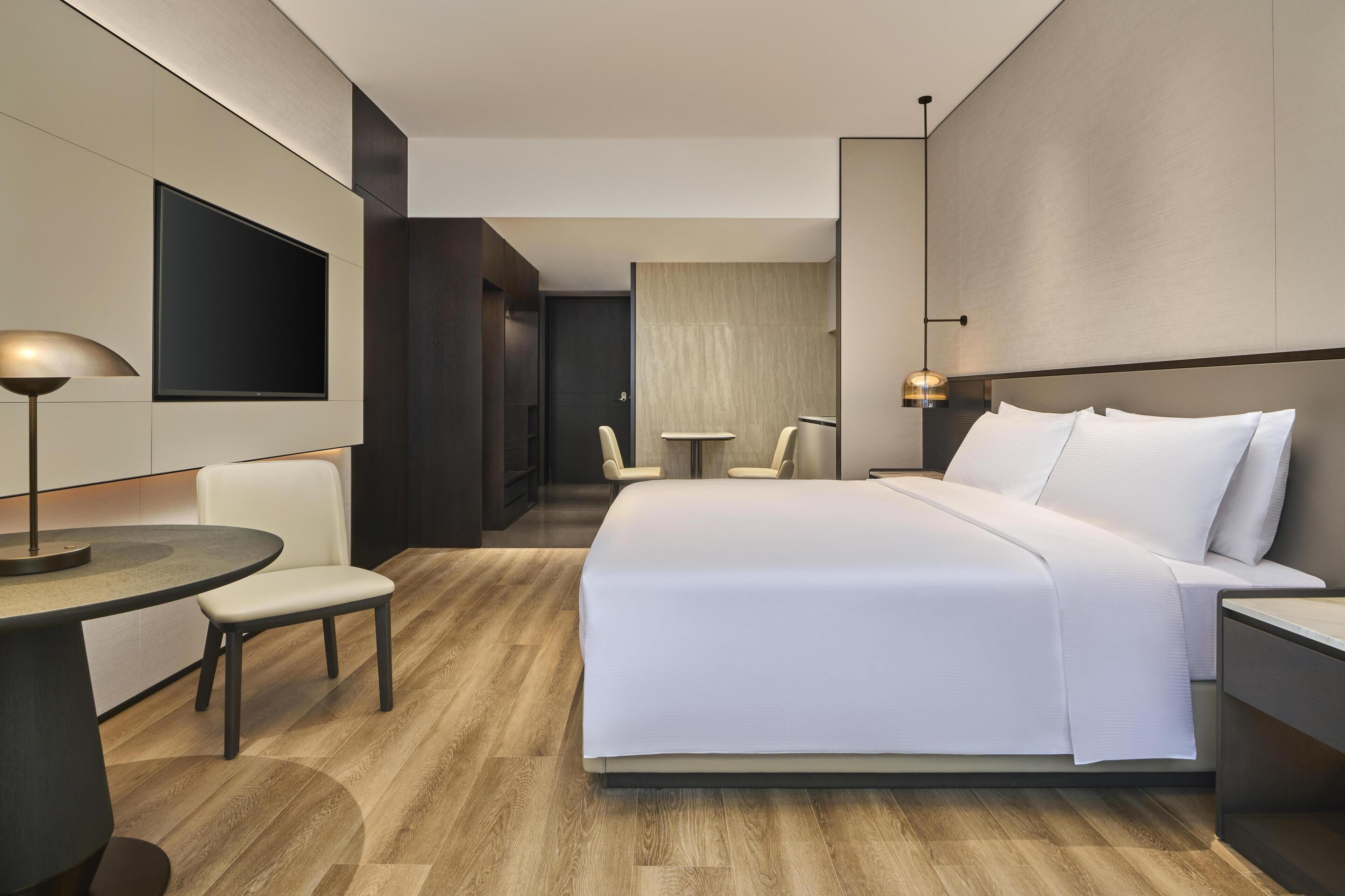 Quarto de hotel moderno com uma cama grande, uma escrivaninha, duas cadeiras, uma TV fixada na parede e uma mesinha.