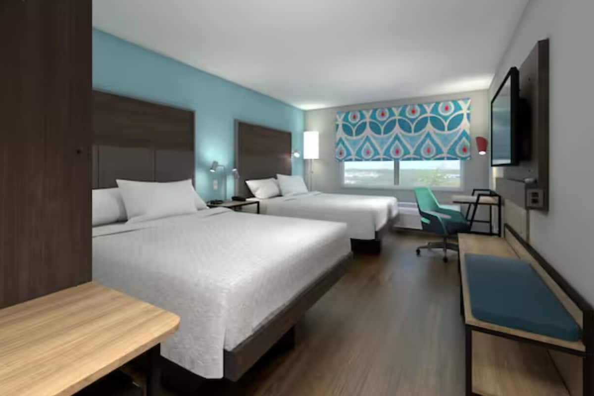 Quarto de hotel com duas camas, uma escrivaninha, uma cadeira, uma TV e uma janela com vista.