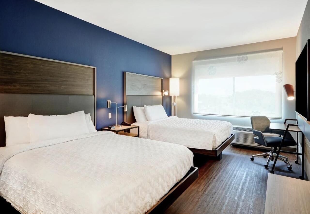 Quarto de hotel com duas camas, uma mesa, uma cadeira e uma janela grande.