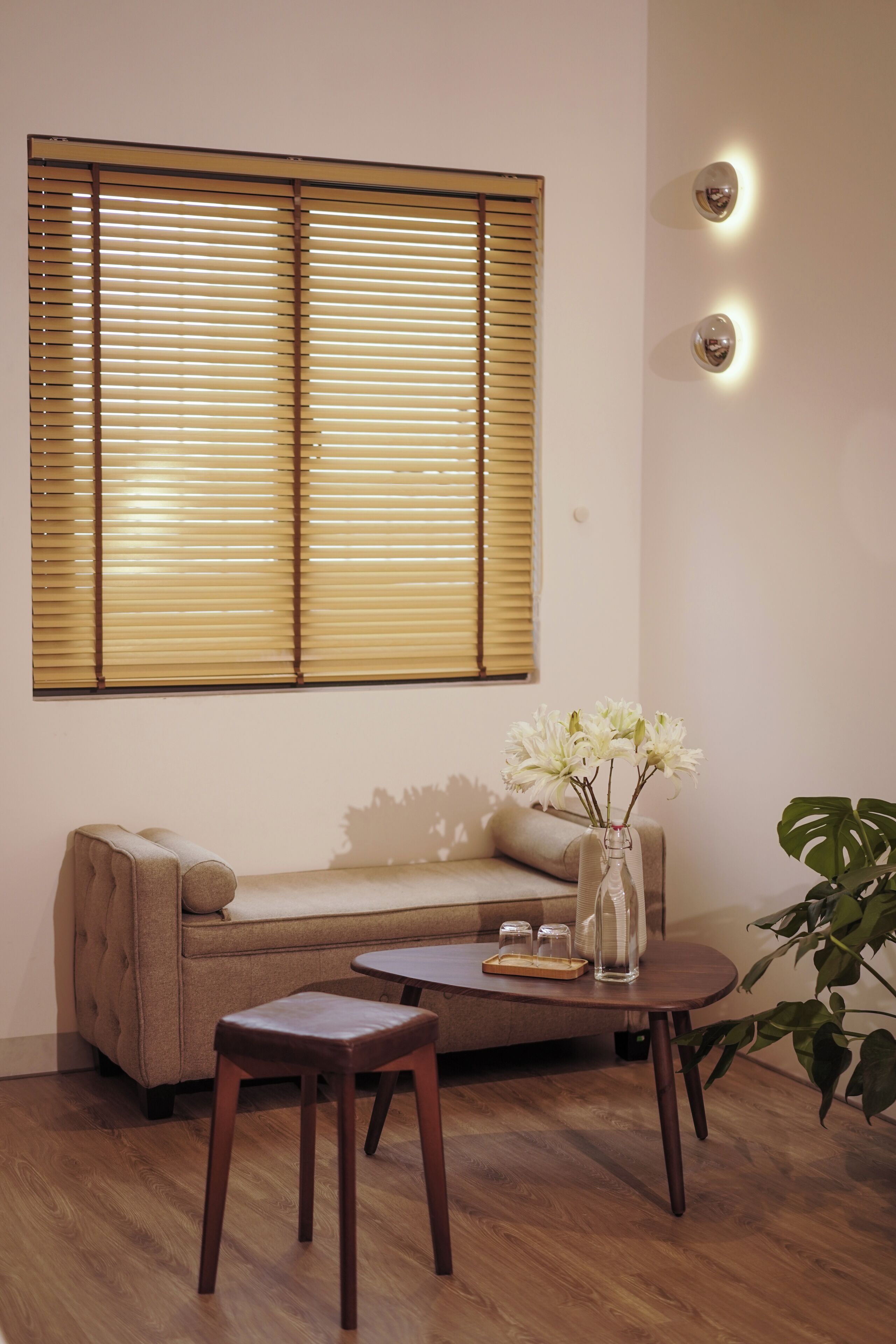 Quarto com sofá, uma mesinha com um vaso de flores, persianas de madeira em uma janela e uma planta em vaso.