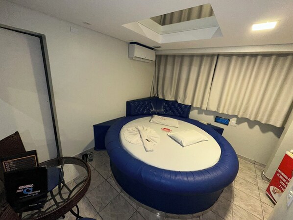 Uma cama redonda com cabeceira e peseira estofadas em azul, colchão branco e luminária no teto.