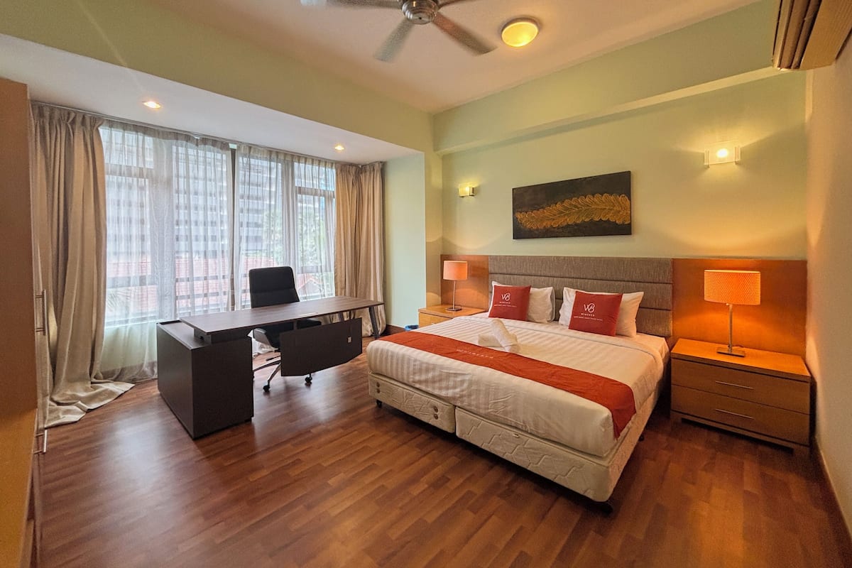 Quarto de hotel com cama, escrivaninha e cadeira. Há um ventilador de teto, uma janela com cortinas e uma luminária na parede.