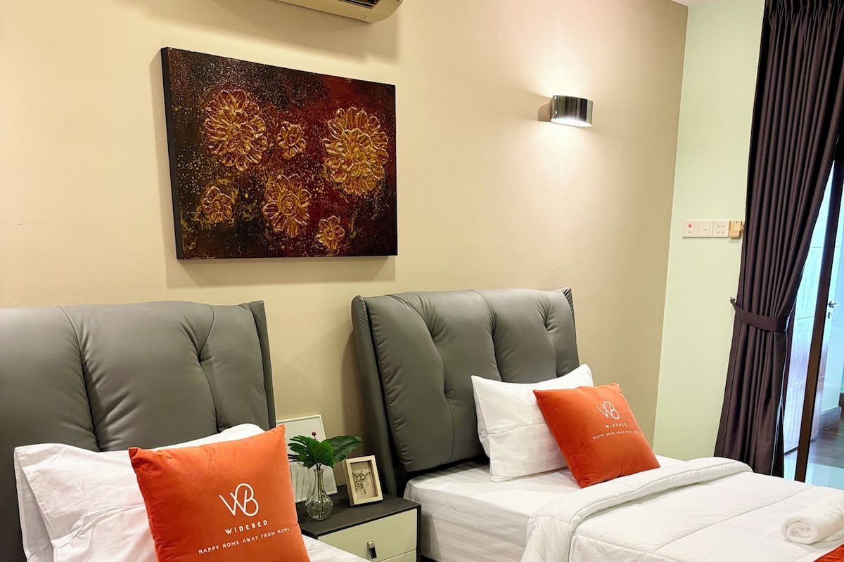 Quarto de hotel com duas camas, cada uma com cabeceira cinza, roupa de cama branca e almofadas laranja. Há um ar condicionado fixado na parede acima das camas, um quadro emoldurado na parede e uma janela com cortinas.