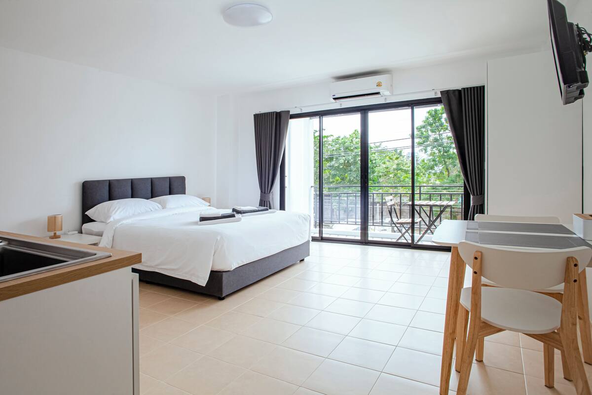 Quarto de hotel moderno com cama, área de refeições com cadeiras, cozinha e varanda com vista para árvores.