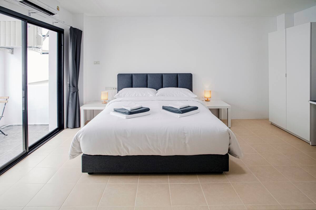 Quarto moderno com uma cama grande, duas mesas de cabeceira com luminárias, uma porta de vidro deslizante para uma varanda e um guarda-roupa.