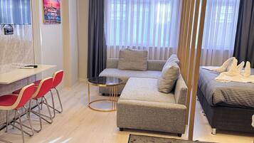 Quarto de hotel moderno com sofá, cama, área de refeições com cadeiras, televisão e uma janela grande com cortinas.