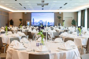 Uma sala de banquetes com mesas redondas preparadas para refeições, com toalhas brancas, copos e centros de mesa florais.