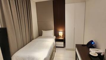 Quarto de hotel com cama, escrivaninha com telefone, ar condicionado, mesa de cabeceira com abajur e armário.