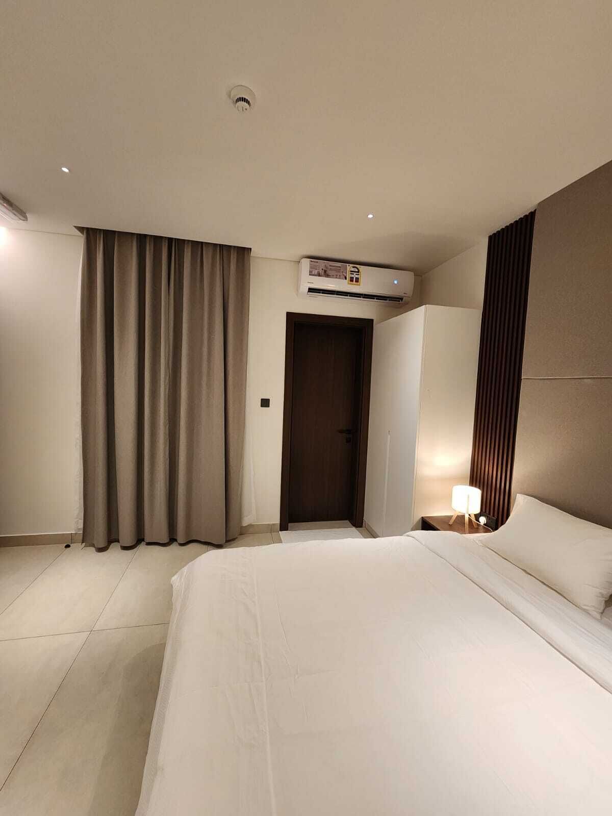 Quarto de hotel com cama, porta, ar condicionado e mesa de cabeceira com abajur.