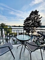 Varanda com mesa e cadeiras de vidro, com vista para a água e uma árvore.