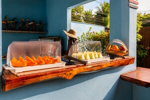Disposição de buffet com uma tábua de madeira, uma tampa de plástico transparente sobre uma travessa de frutas fatiadas e uma cesta de laranjas.