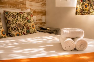 Cama bem arrumada com toalhas brancas dobradas, um travesseiro estampado e uma pequena mesa de cabeceira com controle remoto.