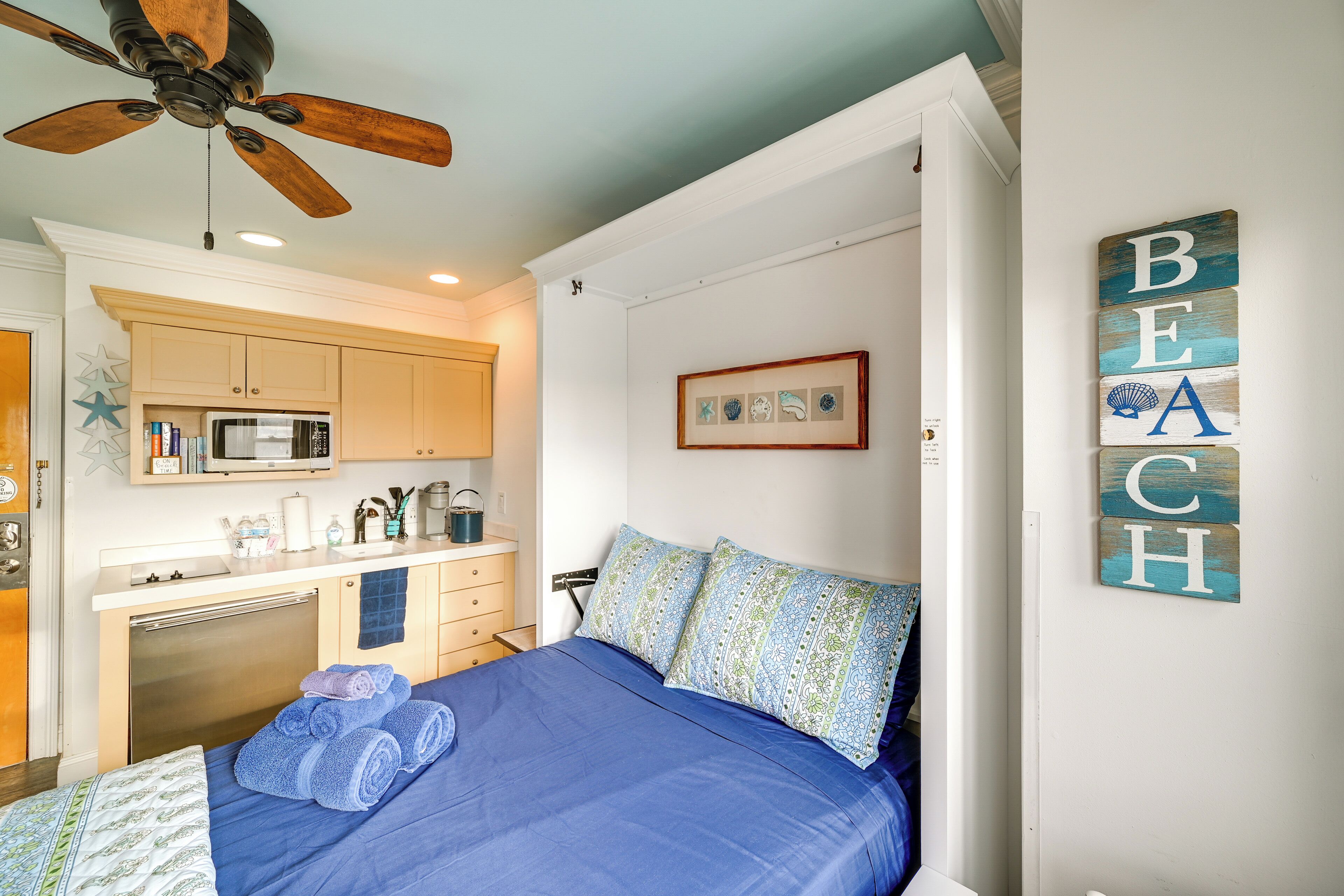 Quarto com cama, ventoinha no teto, cozinha compacta e uma parede com a inscrição 'BEACH'.