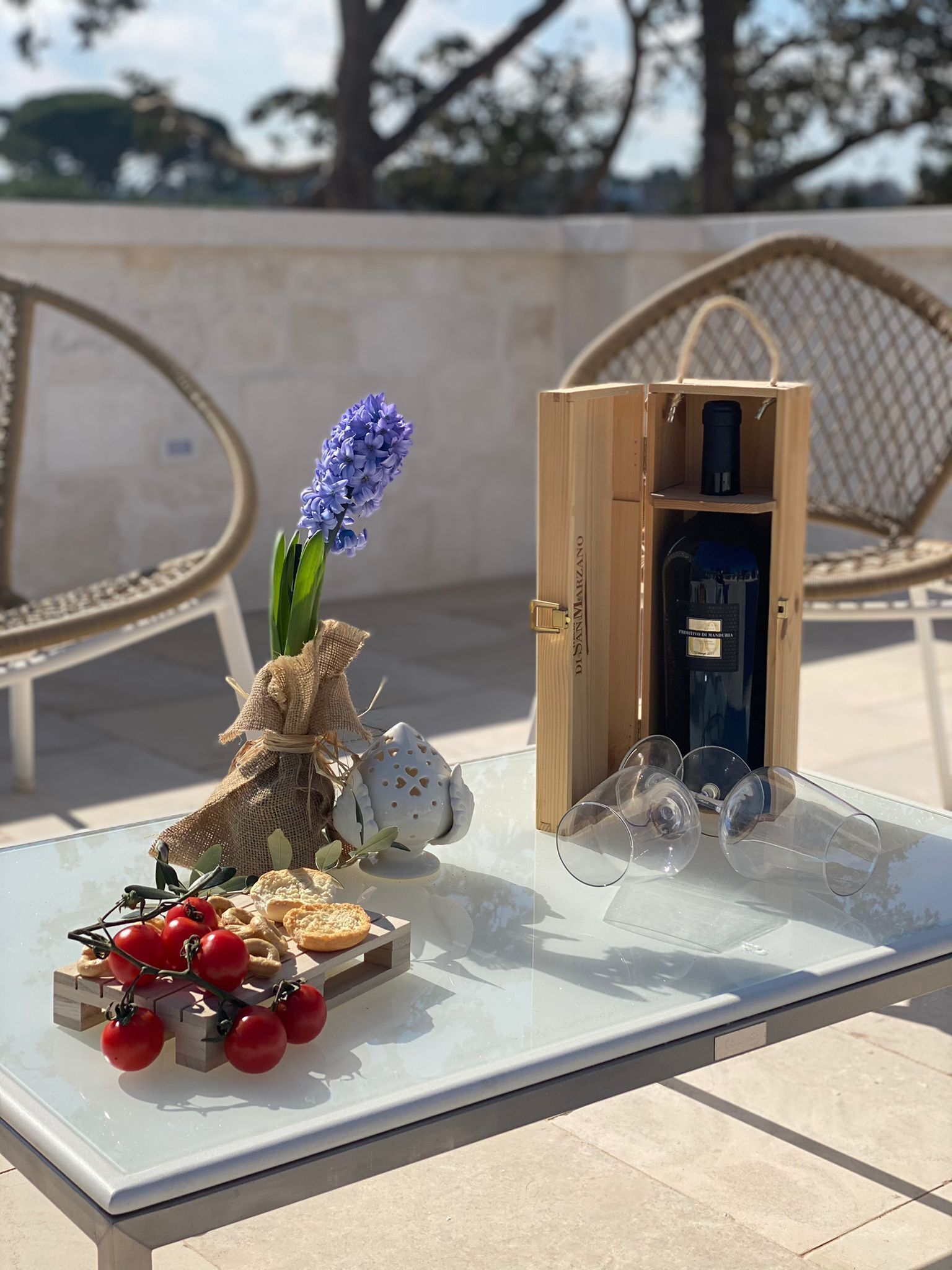 Mesa de vidro com uma garrafa de vinho, copos e uma composição decorativa com tomates vermelhos, uma flor azul e um sapo de cerâmica.