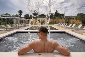 Um homem relaxa em uma piscina, com água salpicando ao redor, cercado por espreguiçadeiras e um pérgola.