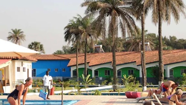 Uma área de piscina com palmeiras, um edifício azul e verde, e pessoas relaxando à beira da piscina.