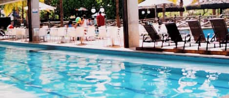 Piscina com espreguiçadeiras e guarda-sóis, cercada por palmeiras e uma área coberta.