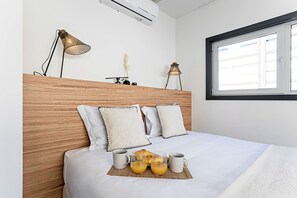 Cama bem arrumada com roupa de cama e almofadas brancas, um tabuleiro com sumo de laranja e chávenas de café, e duas lâmpadas na mesa de cabeceira.